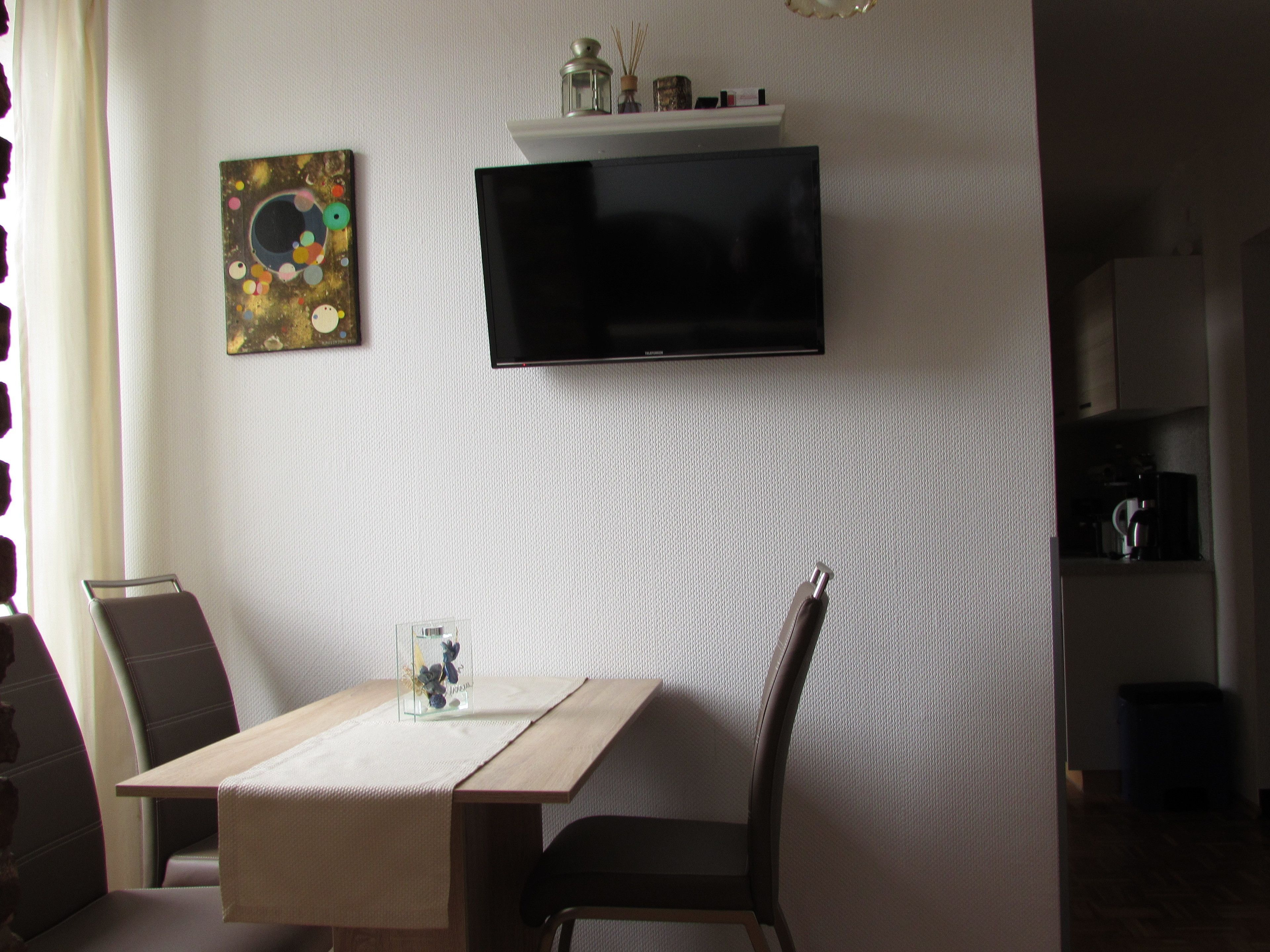 Quarto com televisão de tela plana na parede, uma mesa de jantar com cadeiras e um pequeno objeto decorativo sobre a mesa.