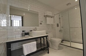 Um banheiro moderno com chuveiro revestido de azulejos brancos, área de banho com box de vidro, vaso sanitário branco e toucador com pia e espelho.