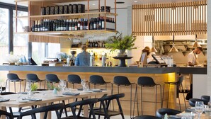 Restaurante moderno com área de bar, prateleiras de madeira com garrafas de vinho e espaço de estar com cadeiras e mesas pretas.