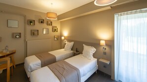 Quarto de hotel com duas camas de solteiro, uma escrivaninha, uma cadeira, uma mesinha, um criado-mudo, um abajur, um espelho e uma janela com cortinas.