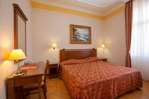 Quarto de hotel com cama, escrivaninha com luminária, cadeira, quadro na parede e janela com cortinas.