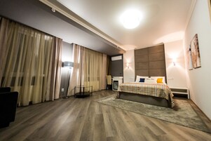 Quarto de hotel com cama, escrivaninha e cadeira. Possui uma janela com cortinas, uma luminária na parede e iluminação no teto.