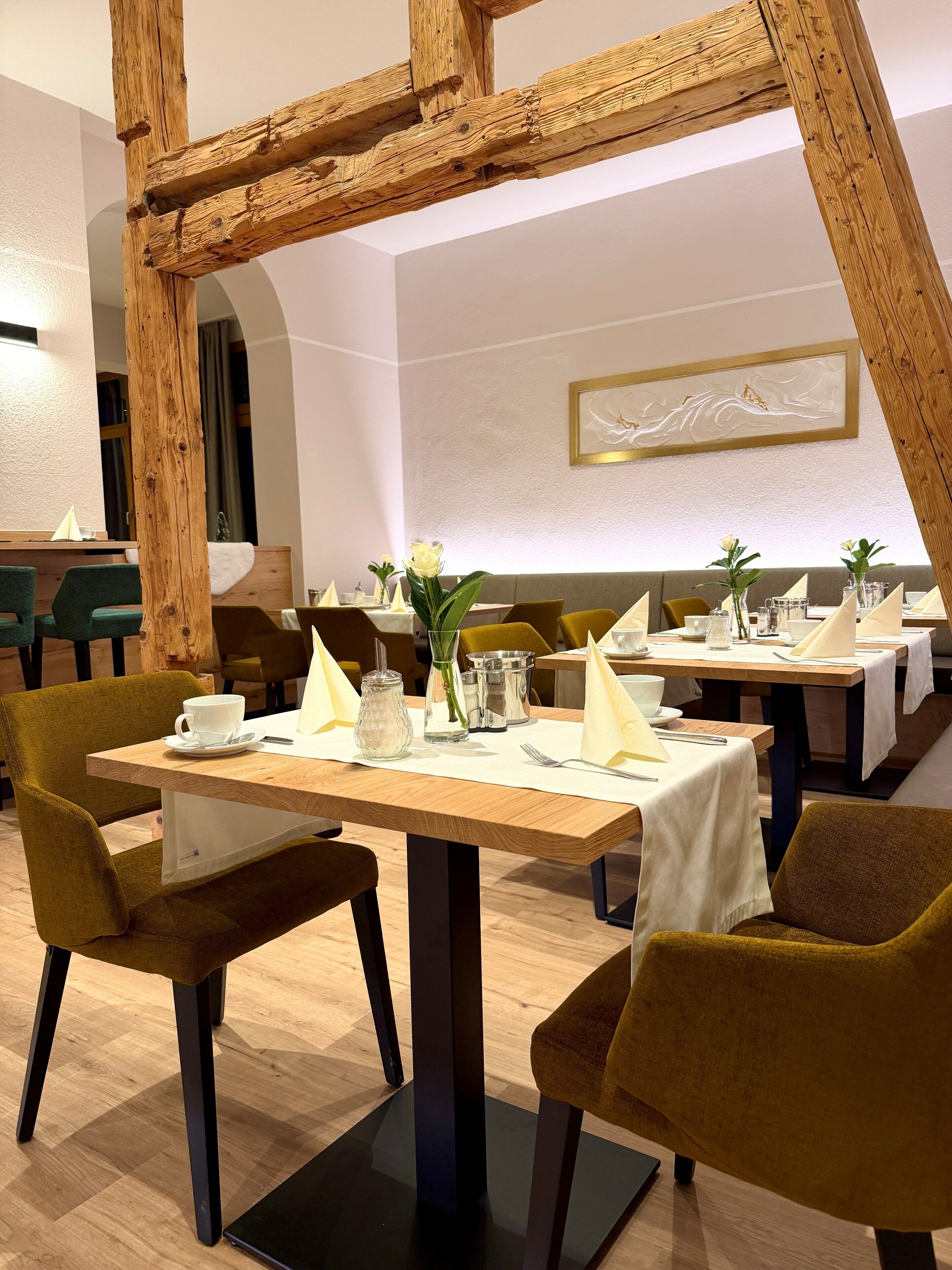 Restaurante com vigas de madeira, cadeiras estofadas verdes e mesas dispostas com toalhas e guardanapos brancos.