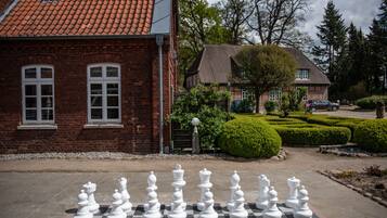 Um tabuleiro de xadrez grande montado ao ar livre em um jardim, com um edifício de tijolos ao fundo.