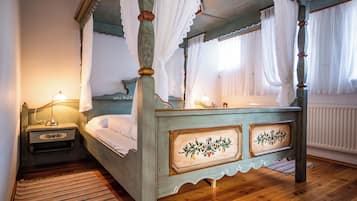 Uma cama de dossel com estrutura de madeira e painéis florais, uma mesa de cabeceira com abajur e uma janela com cortinas translúcidas.