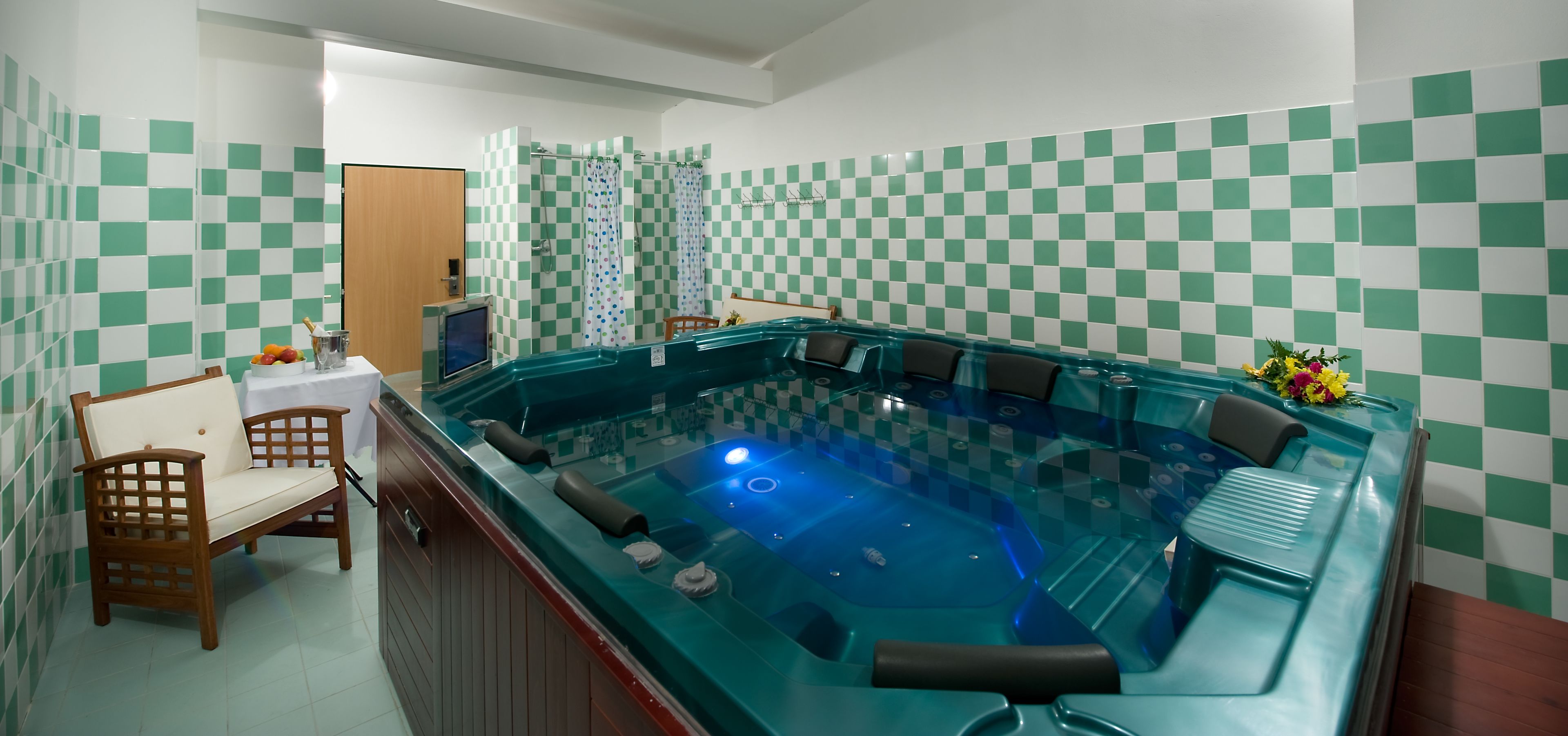 Área de spa com parede revestida de azulejos brancos e verdes em padrão xadrez, uma banheira de hidromassagem em vidro, cadeiras de madeira e uma pequena mesa com televisão e frutas.