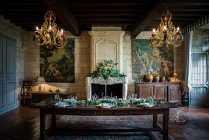 Uma sala de jantar com uma grande mesa de madeira posta para refeição, ladeada por duas luminárias ornamentadas, uma lareira com arranjo floral e uma parede de pedra com um mural.