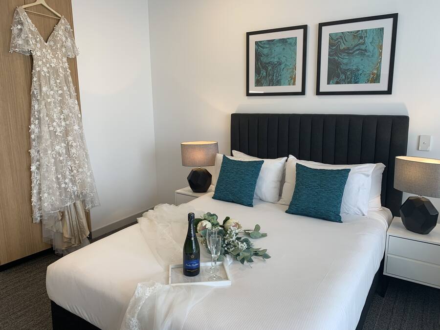Quarto de hotel com uma cama, duas pinturas abstratas emolduradas, uma mesa de cabeceira com abajur, uma garrafa de champanhe e um arranjo de flores.