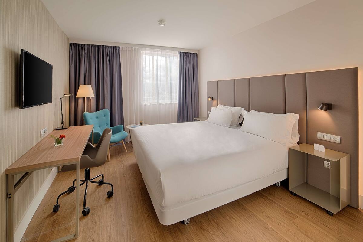 Quarto de hotel com uma cama grande, uma escrivaninha com cadeira, televisão e luminária.