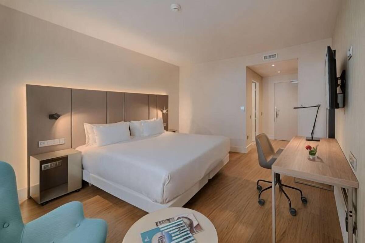 Quarto de hotel com uma cama grande, mesas de cabeceira, uma escrivaninha com cadeira, uma televisão e uma pequena mesa com um vaso.
