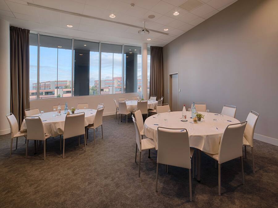Sala de conferências com mesas redondas, cadeiras e grandes janelas que oferecem vista para prédios.