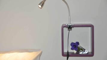 Espelho de moldura quadrada com um aparelho de luz curvo acima, refletindo uma flor roxa num recipiente de vidro.