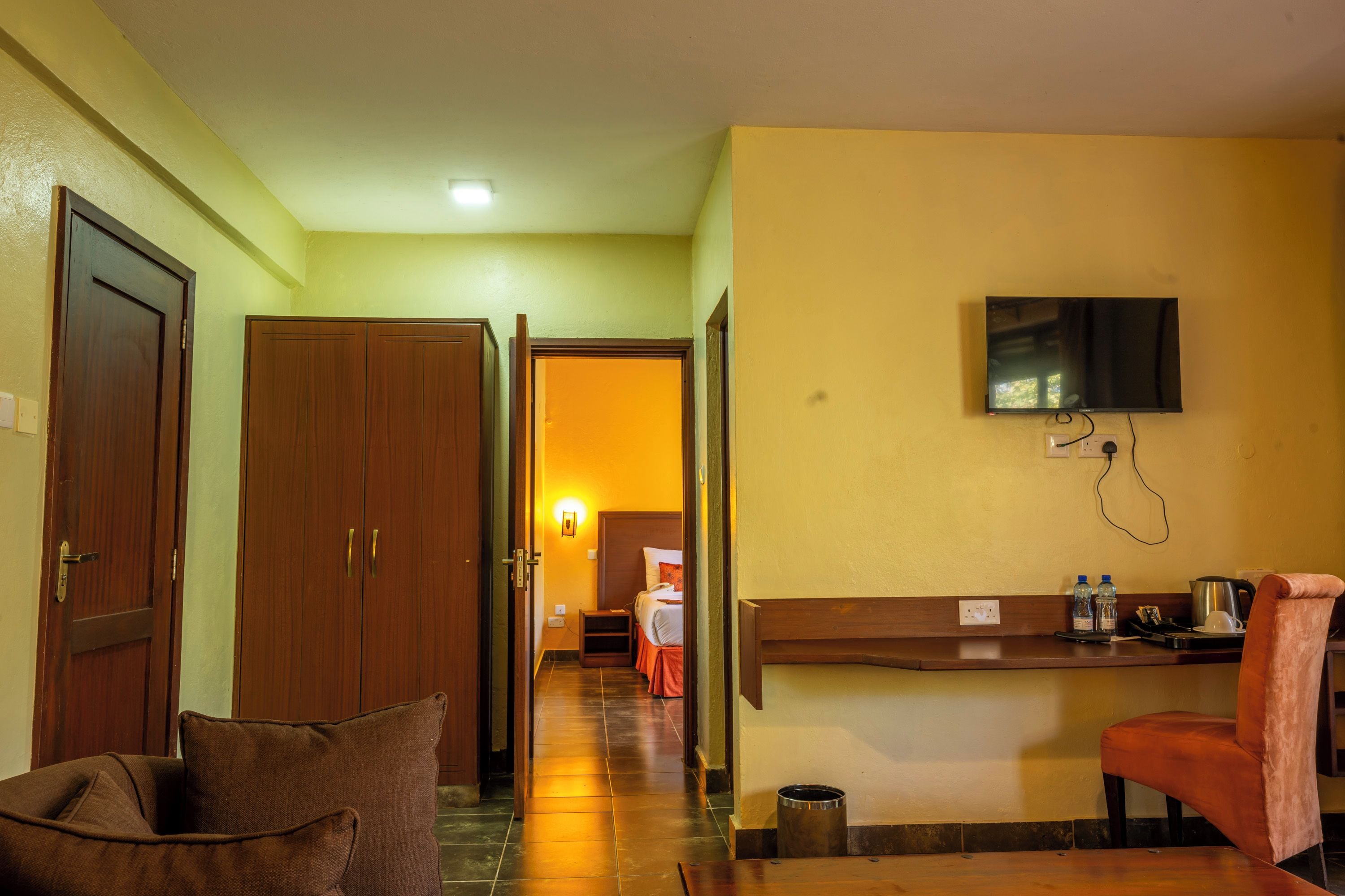 Quarto de hotel com armário de madeira, cama com saia vermelha, televisão na parede e pequena mesa com chaleira e xícaras.