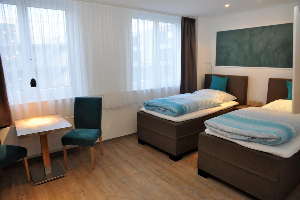 Quarto de hotel com duas camas, uma mesa pequena, uma cadeira e uma janela com cortinas finas.