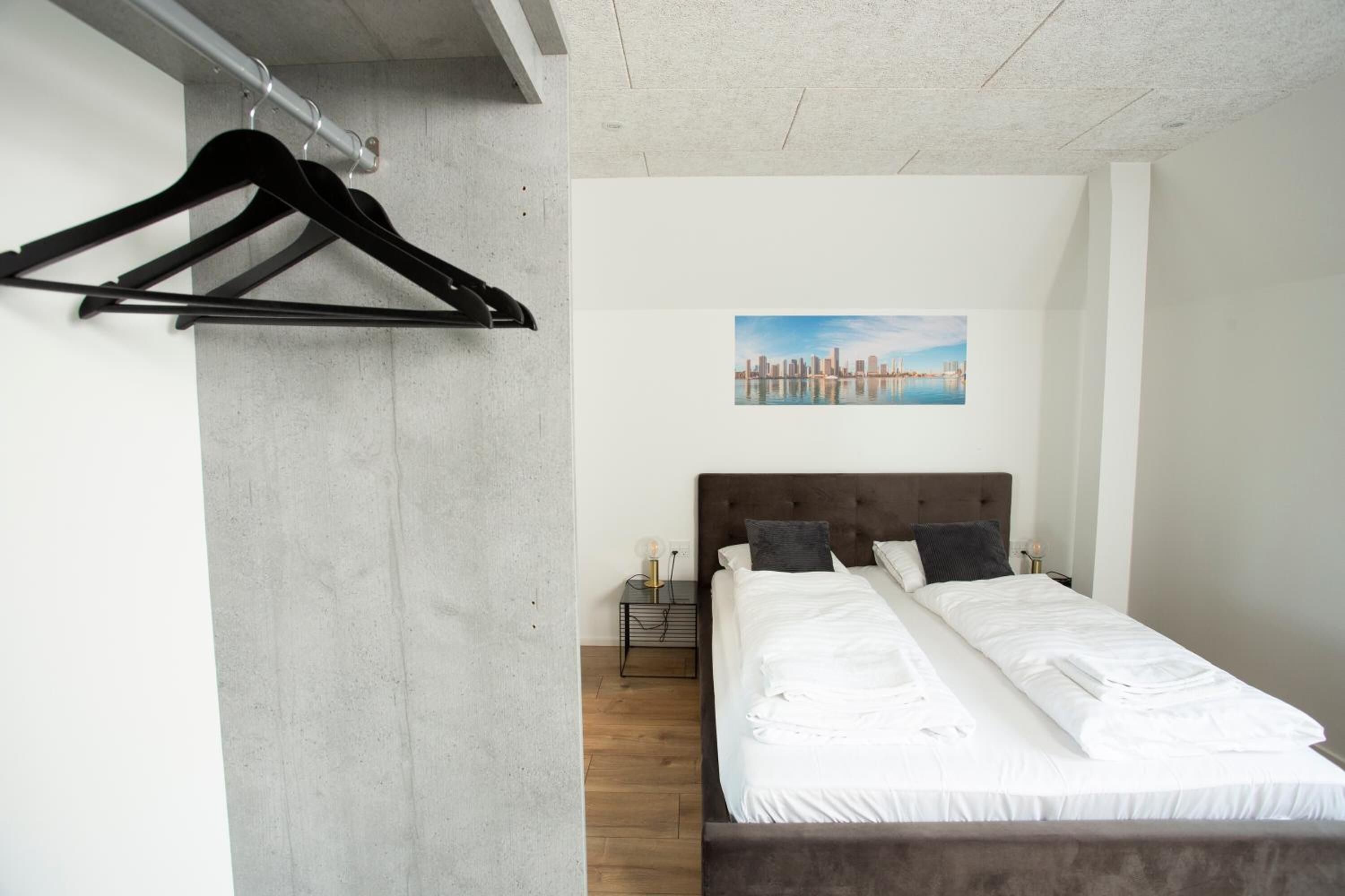 Quarto de hotel moderno com uma cama grande, cabeceira, cabideiro fixado na parede e uma pintura de paisagem urbana.