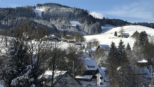 Uma aldeia coberta de neve com casas tradicionais, uma pista de esqui e uma colina arborizada.
