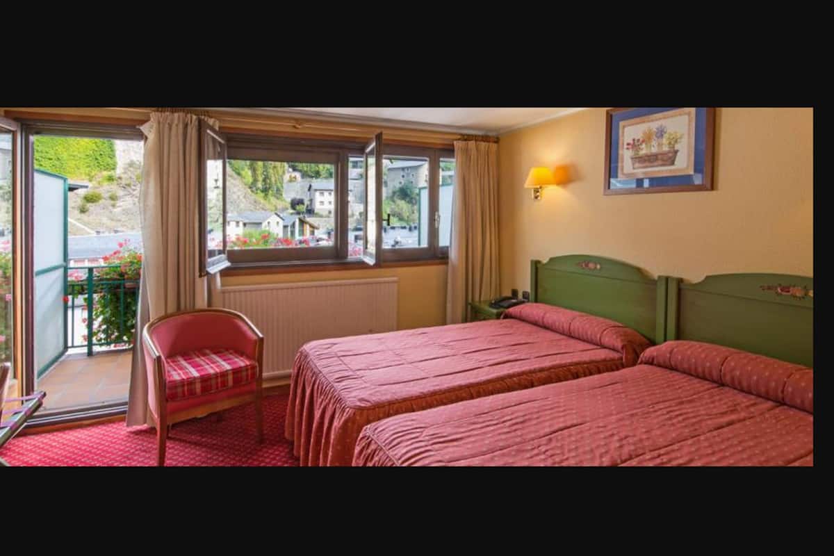 Quarto de hotel com duas camas, uma cadeira, janela com vista para o exterior e uma luminária na parede.