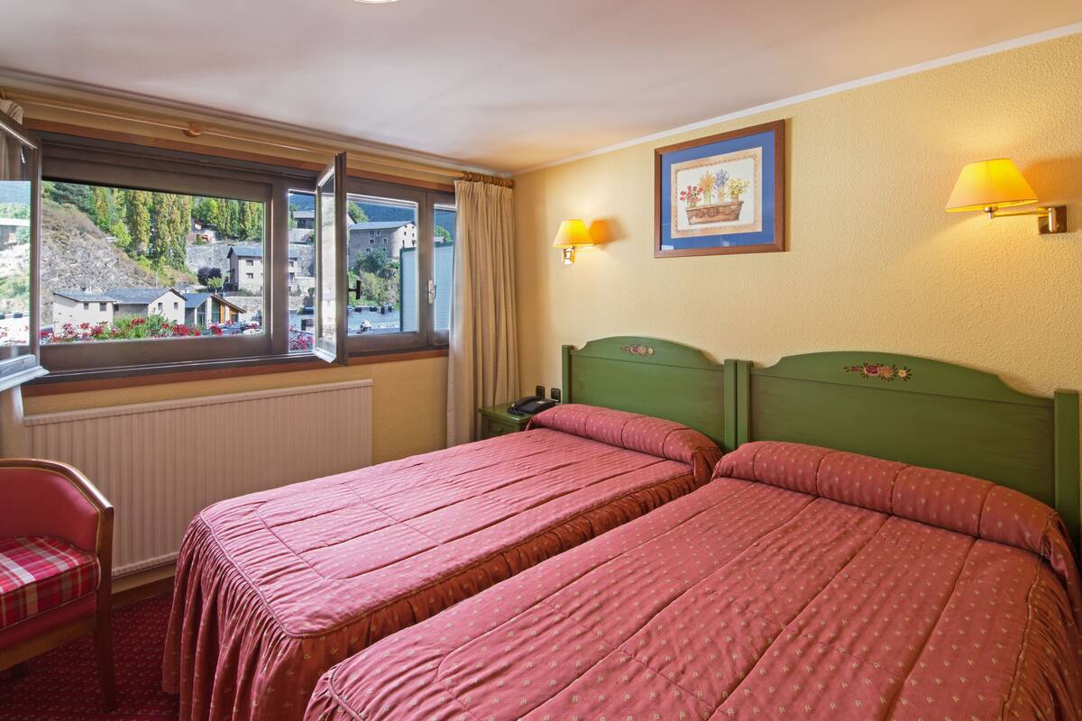 Quarto de hotel com duas camas, janela com vista para a aldeia, uma cadeira e uma mesa de cabeceira com abajur.