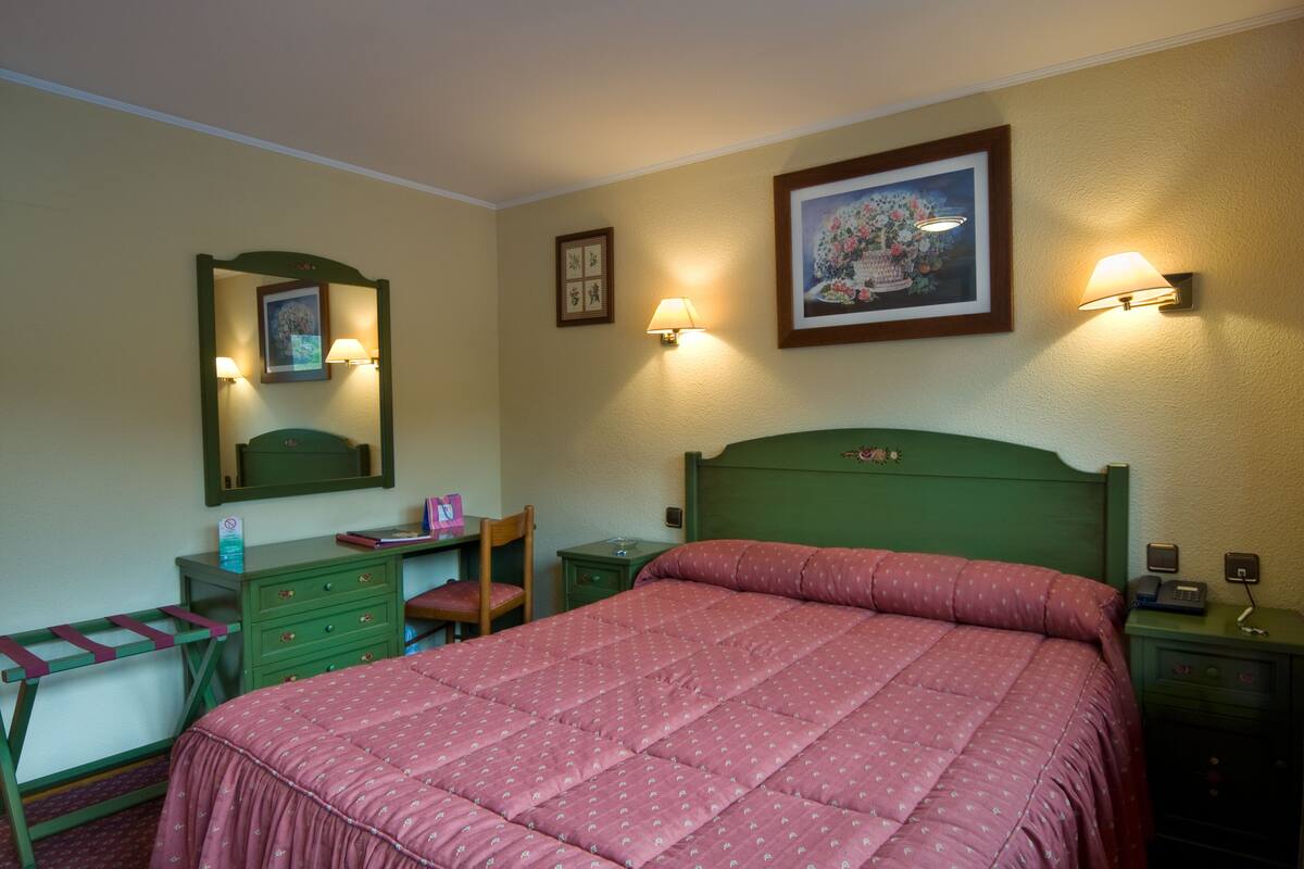 Quarto de hotel com cama verde, escrivaninha com cadeira, cômoda e mesa de cabeceira com abajur.