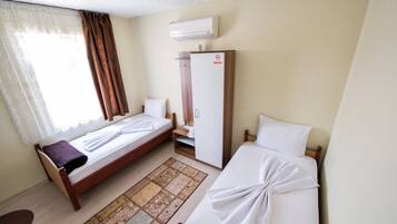 Quarto pequeno e limpo com duas camas de solteiro, um guarda-roupa de madeira, banheiro com chuveiro e janela com cortinas.