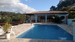 Área da piscina com um alpendre coberto, piso de pedra e vista para uma encosta.