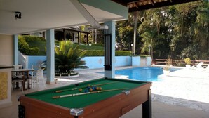 Mesa de bilhar com bolas, mesa de sinuca e piscina numa área externa coberta com vista para um jardim ajardinado e piscina.