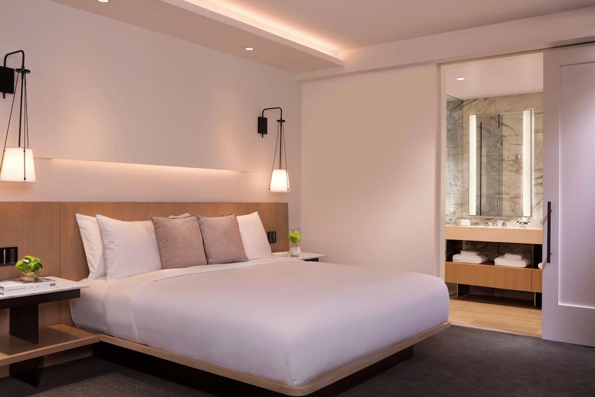 Quarto moderno com cama grande, mesas de cabeceira, um criado-mudo, luminária na parede e banheiro com chuveiro e pia de mármore.