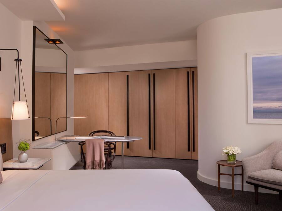 Quarto de hotel moderno com cama, escrivaninha, cadeira, espelho e pintura de paisagem.