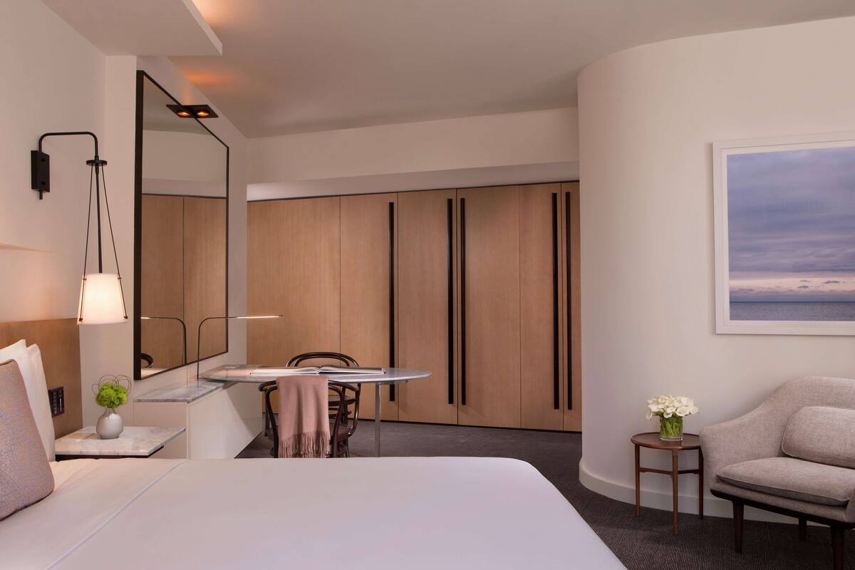 Quarto de hotel moderno com uma cama grande, uma escrivaninha, uma cadeira, uma mesinha com flores, um espelho e vista para o mar.