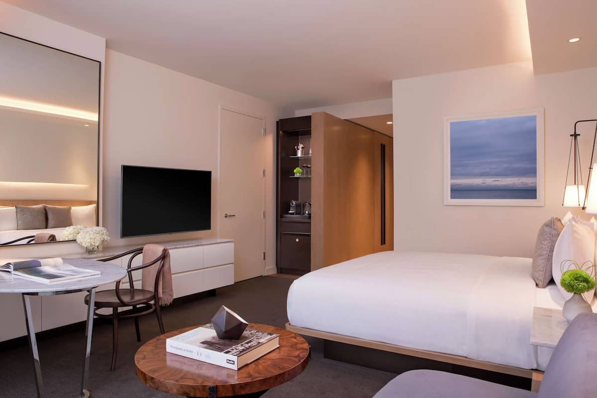 Quarto de hotel moderno com uma cama grande, televisão de tela plana, escrivaninha com cadeira e uma mesinha redonda.