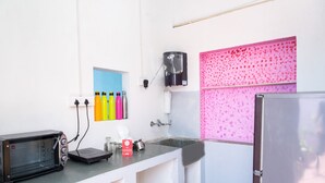 Cozinha compacta com micro-ondas, torradeira e geladeira. Há garrafas de água coloridas e uma cafeteira no balcão.