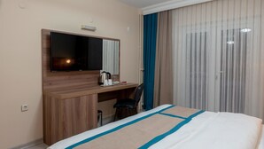 Quarto de hotel com cama, escrivaninha, cadeira, televisão e janela com cortinas.
