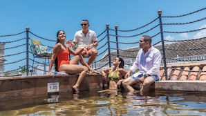 Um grupo de pessoas sentadas à beira da piscina, com um céu claro ao fundo.