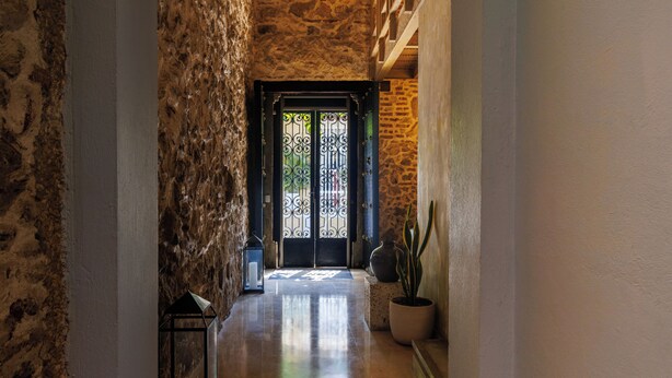 Um corredor com parede de pedra, uma porta preta com detalhes em vidro decorativo, um vaso de planta e um candeeiro.