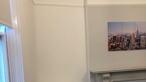 Um quarto com cômoda branca, pintura de paisagem urbana na parede e janela com cortinas.