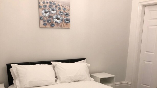 Cama bem arrumada com roupa de cama branca e duas toalhas castanhas por cima, uma mesinha de cabeceira branca com uma luminária, e um quadro na parede.