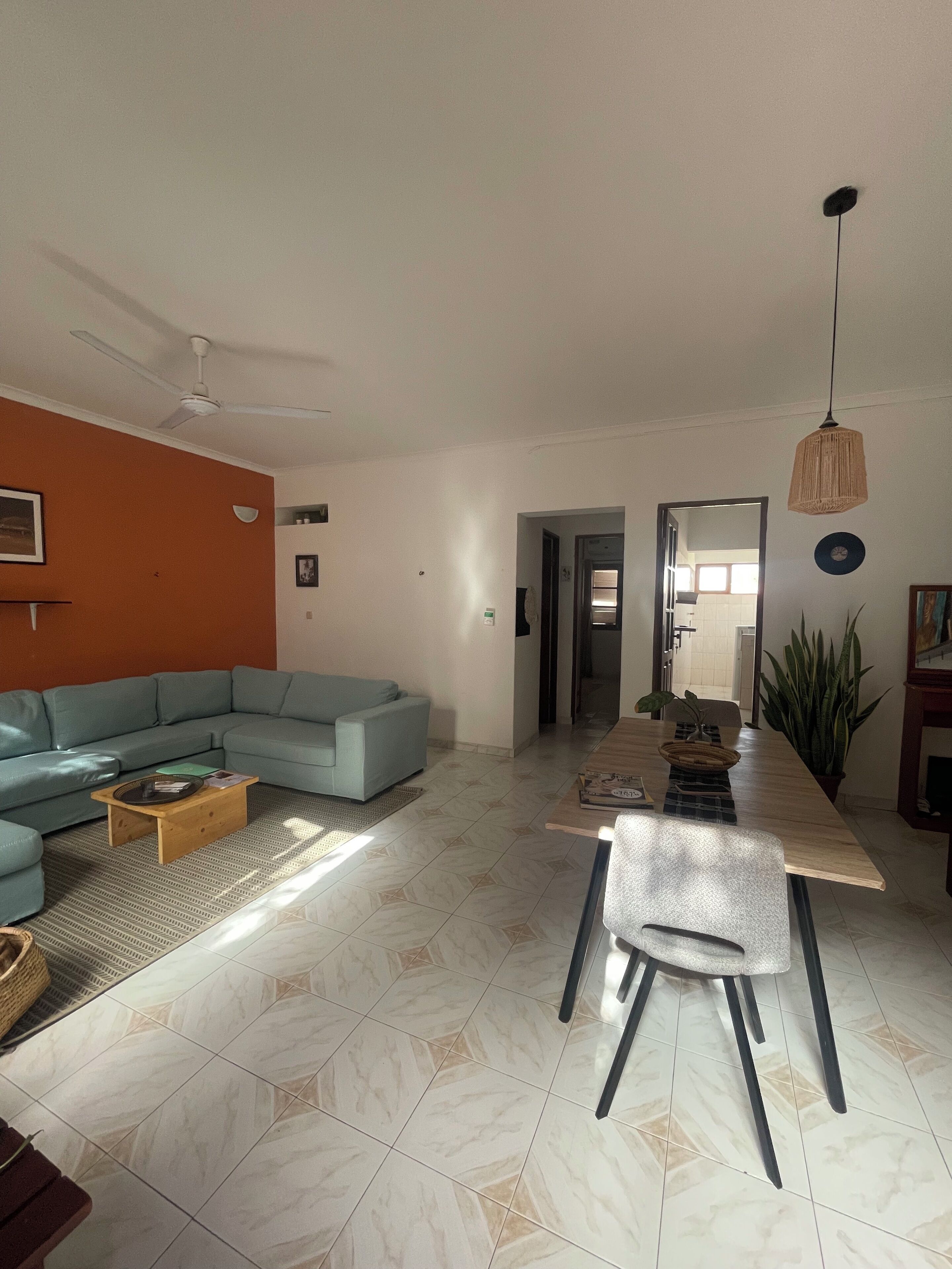 Uma sala de estar com sofá, mesa de centro e mesa de jantar com cadeiras. Há um ventilador de teto, um lustre e um vaso de planta.