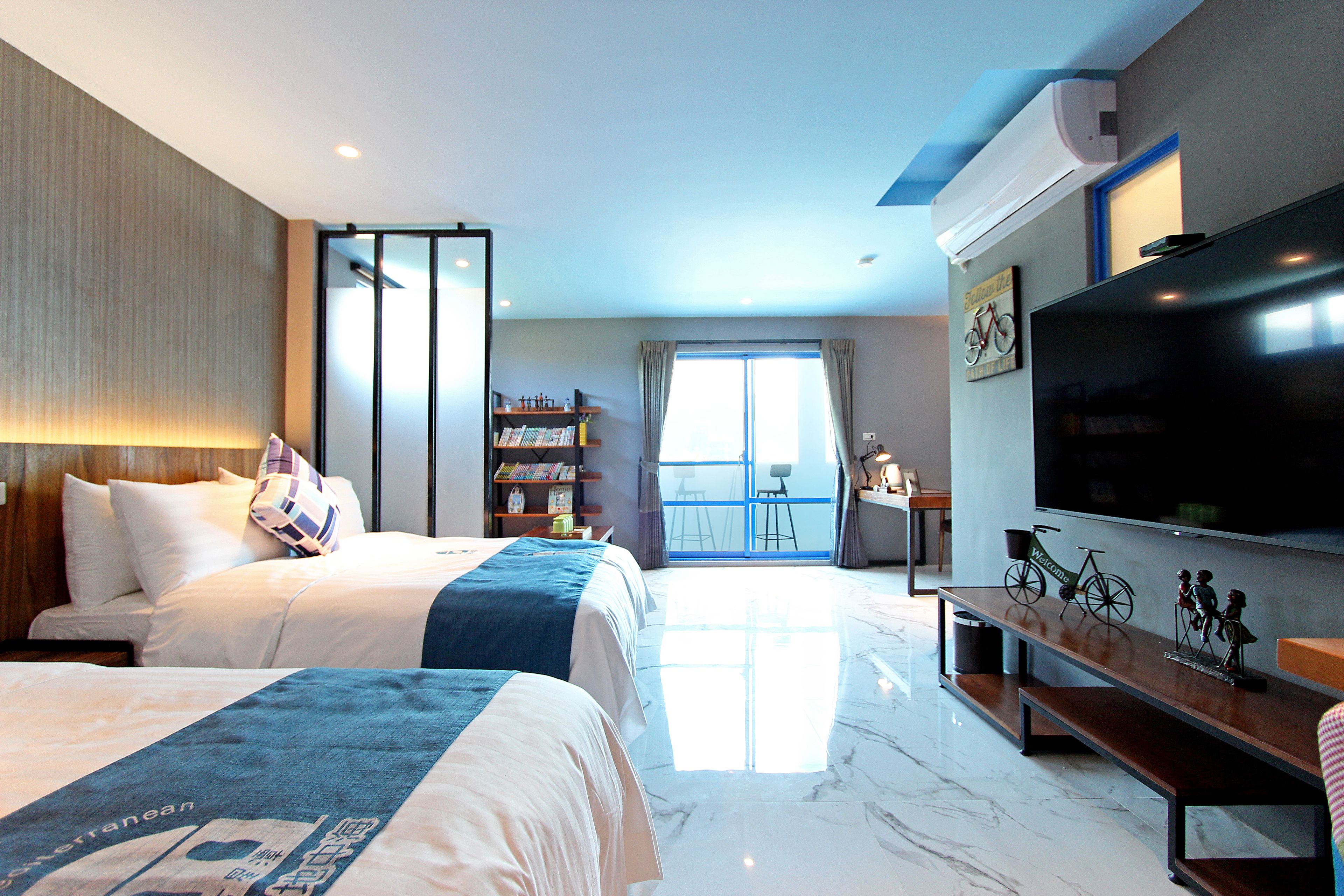 Quarto de hotel com duas camas, uma televisão grande, uma estante e vista para o exterior.