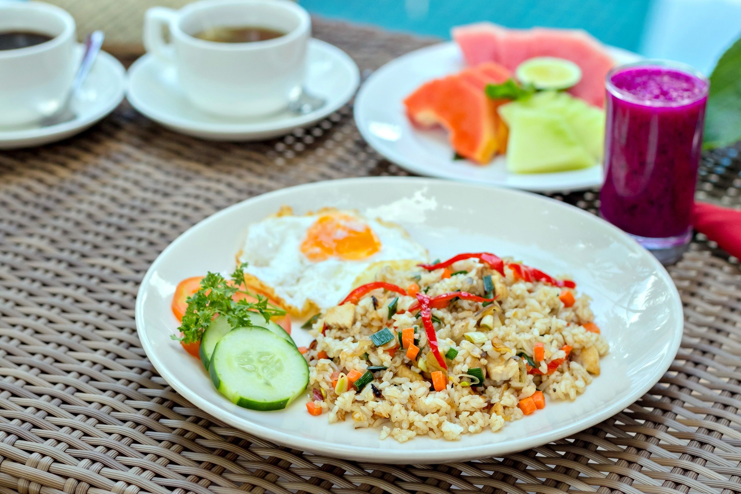 Um prato de arroz frito com legumes e um ovo, um prato de melancia e melão, e um copo de smoothie roxo sobre uma mesa de palha.