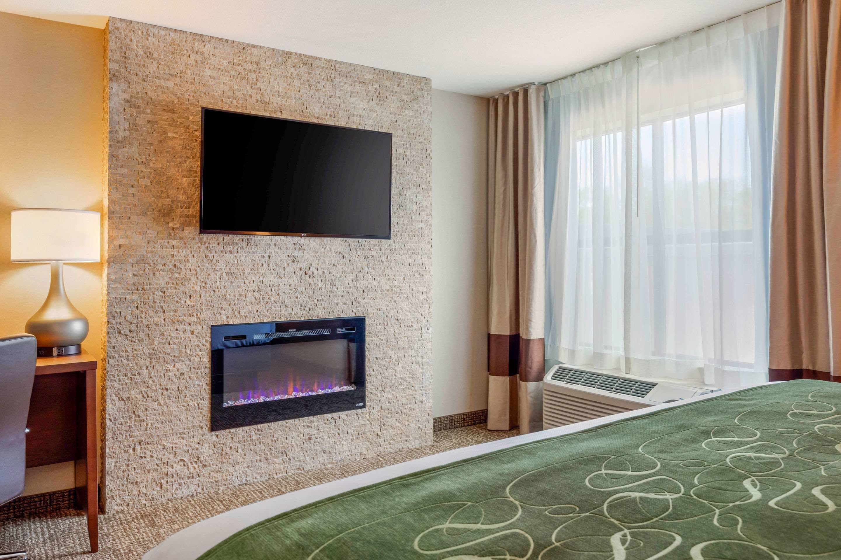 Quarto de hotel com televisão de tela plana fixada em parede texturizada, lareira embutida, cama com colcha estampada em verde, escrivaninha com abajur e grande janela com cortinas leves.
