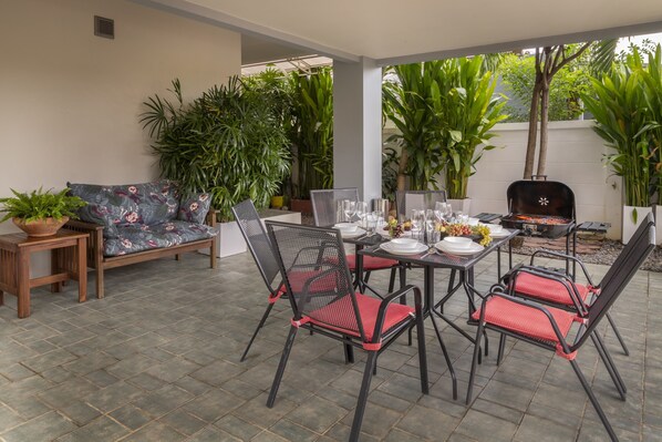 Um pátio externo com uma mesa de jantar posta para quatro pessoas, rodeada por cadeiras com almofadas vermelhas. Há um banco de madeira com uma almofada floral, e vasos de plantas estão dispostos pela área.