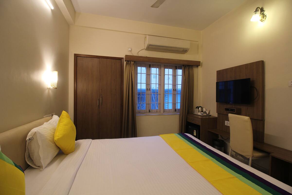 Quarto de hotel com cama, escrivaninha, cadeira, televisão e janela com cortinas.