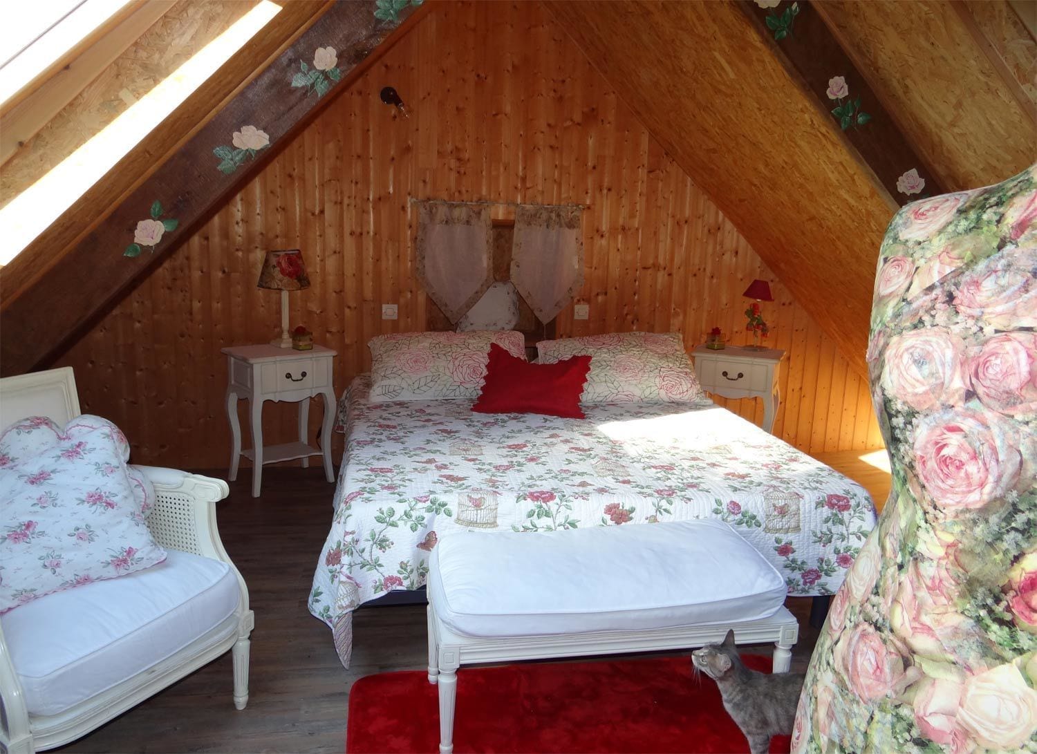 Um quarto de sótão acolhedor com teto inclinado, paredes de madeira, colcha estampada com flores, travesseiro vermelho, cadeira branca e um banquinho vermelho.