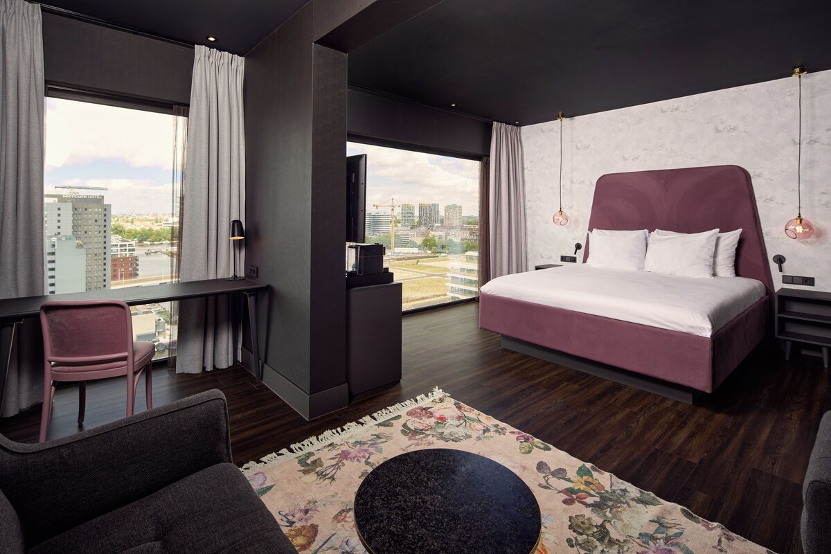 Quarto de hotel moderno com uma cama grande, uma escrivaninha, uma cadeira e vista para a cidade.
