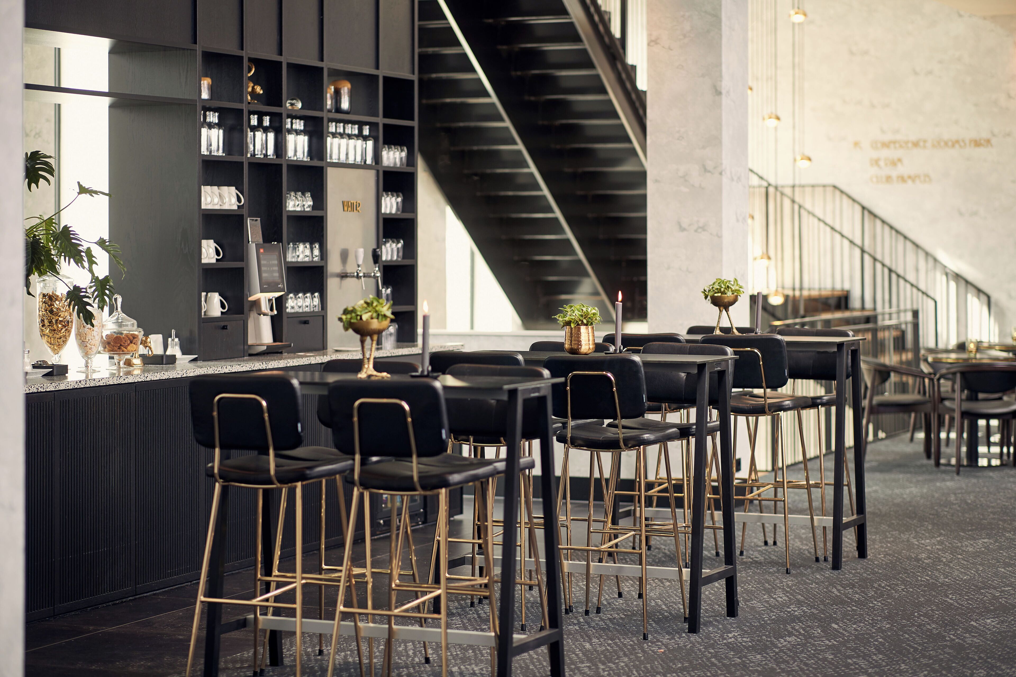 Uma área de bar moderna com banquetas pretas, um balcão com copos e uma escada ao fundo.