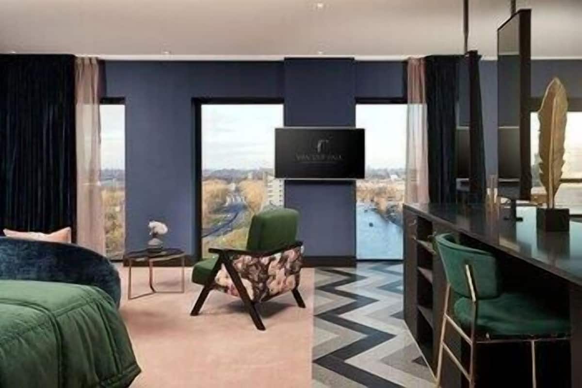 Quarto de hotel com cama, poltrona, mesinha, televisão e uma janela grande com vista para a cidade.