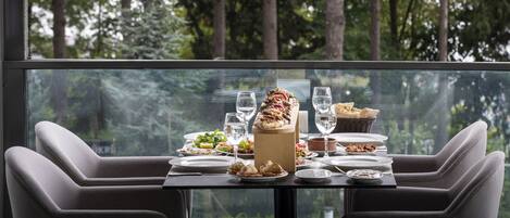 Mesa posta com pratos, copos e tábua de madeira com comida, vista através de uma janela de vidro com um panorama de floresta ao fundo.