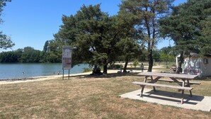Um parque à beira do lago com mesas de piquenique, uma placa de vigilância e um pequeno abrigo.