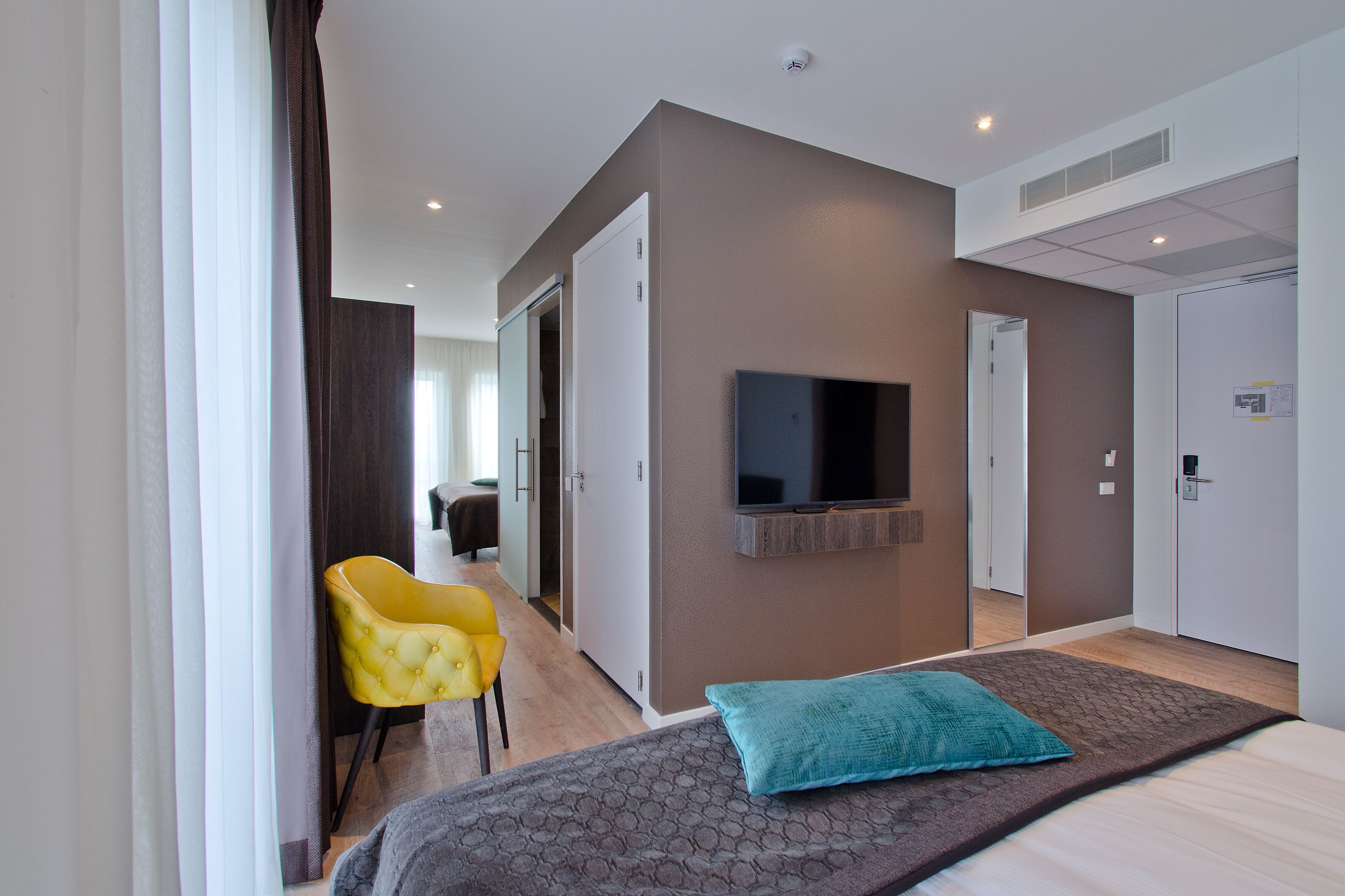 Quarto de hotel moderno com uma cama, televisão fixada na parede, uma cadeira amarela e uma janela grande.