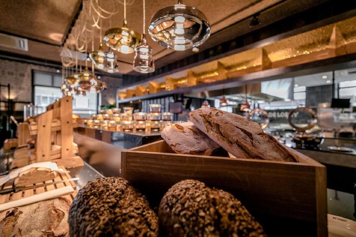 Vitrine de padaria com diversos pães e doces, caixas de madeira, luminárias pendentes e parede interna de tijolos.